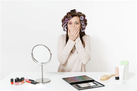 흰색 바탕에 격리 설정된 얼굴 장식 화장품으로 메이크업을 적용 하는 테이블에 앉아 손으로 Curlers 커버 입으로 재미 있는 여자 뷰티 여성 패션 라이프 스타일 개념입니다