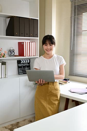 한 여자가 노트북과 컴퓨터 모니터를 들고 책상 앞에 서 있습니다 그녀는 미소를 지으며 손에 노트북을 들고 있습니다 책상에는 책