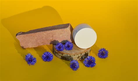 화장품을위한 흰색 플라스틱 항아리가 노란색 배경에 파란색 옥수수 꽃이있는 나무 연단에 서 있습니다 0명에 대한 스톡 사진 및