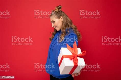 빨간 리본으로 흰색 선물 상자를 들고 파란색 트렌디 한 후드를 입고 빨간색 배경 벽을 통해 고립 된 매력적인 젊은 금발 여성의 샷 놀라움에 대한 스톡 사진 및 기타 이미지