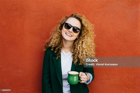주황색 스튜디오 벽에 포즈를 취하는 카메라에 미소를 지으며 카푸치노 컵을 들고 선글라스와 재킷을 입고 곱슬 머리를 한 예쁜
