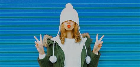 파란 배경에 폼 폼이 달린 흰색 모자를 쓰고 달콤한 공기 키스를 보내는 입술을 불고 있는 젊은 여성의 겨울 초상화 20 29세에