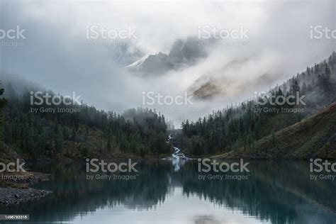 산 개울은 숲 언덕에서 빙하 호수로 흐릅니다 안개 낀 눈의 바위 산 작은 강과 침엽수 림이 잔잔한 고산 호수에 반영되었습니다 이른 아침에 고요한 안개가 자욱한 풍경 대기감에