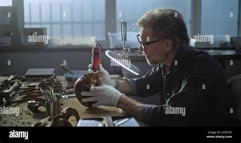 Mature Archaeologist In Gloves Examines Prehistoric Human Skull Under Magnifying Lamp In