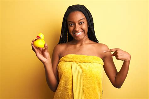 자신에게 손가락을 가리키는 깜짝 얼굴과 고립 된 노란색 배경을 통해 장난감 오리를 들고 샤워 수건을 입고 아프리카 계 미국인