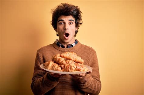 고립 된 노란색 배경 위에 서있는 크루아상과 접시를 들고있는 젊은 잘생긴 남자는 깜짝 얼굴로 충격에 무서워하고 두려움과 두려움 표현으로 흥분했습니다 놀라움에 대한 스톡 사진
