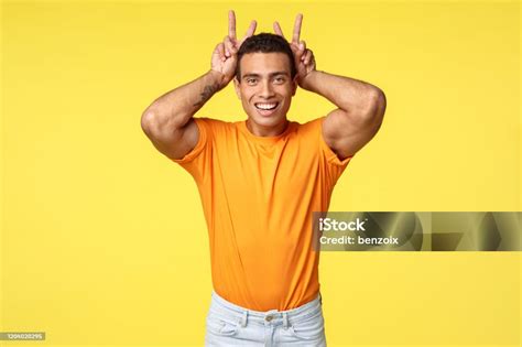 오렌지 색 티셔츠에 귀여운 남성과 바보 히스패닉 남자 친구 평화 제스처를 머리 뒤에 토끼 귀를 즐겁게 미소 노란색 배경을 통해 조카와 함께 장난 주위에 장난 토끼를 모방