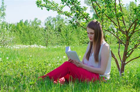 젊은 아주 화창한 날에 꽃이 만발한 사과 나무와 읽기 책 아래 잔디에 앉아 흰 상단과 빨간 바지에 여자 텍스트에 대 한 공간을 복사 개화기에 대한 스톡 사진 및 기타 이미지