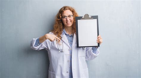 그런 지 회색 벽 표시 클립보드 깜짝 얼굴 자신을 가리키는 손가락에 빨간 머리 젊은 의사 여자 가리키기에 대한 스톡 사진 및 기타 이미지 Istock