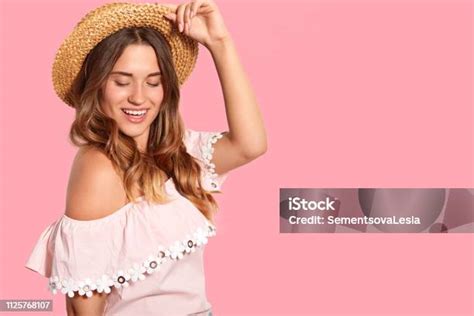 유행 기쁘게 유럽 여자 아래로 집중 밀 짚 모자에 손을 유지 부드러운 미소가 있다 옆으로 텍스트에 대 한 빈 공간을 가진 핑크 배경 위에 절연 세련 된 여름 옷을 입고