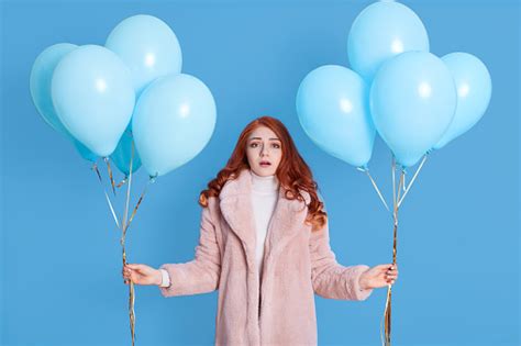젊은 백인 여성은 무서워하고 충격을 보고 입을 열고 카메라를 바라보며 깜짝 놀란 채 헬륨 풍선을 손에 들고 옅은 분홍색 모피