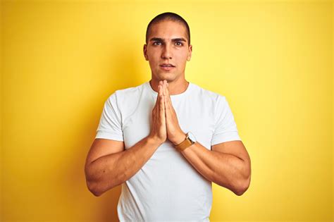 노란색 고립 된 배경에 캐주얼 흰색 티셔츠를 입고 젊은 백인 남자는 매우 감정적이고 걱정 얼굴에 희망 표현과 함께 손으로 구걸하고기도합니다 용서를 구함 종교 개념 T 셔츠에