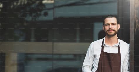 성공적인 중소기업 남성 바리스타 커피숍 유리 벽 앞에 기대어 있다 수염 난 남자의 초상화는 복사 공간이 있는 카메라를 보고 갈색 앞치마를 입고 있다 사진