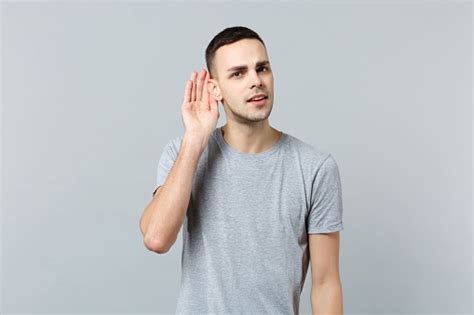 스튜디오에서 회색 벽 배경에 고립 된 귀 근처 손을 잡고 카메라 청력을 찾고 캐주얼 복장에 호기심 젊은 남자의 초상화 사람들은 진심으로 감정 라이프 스타일 개념입니다 복사