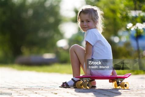 흰색 반바지와 티셔츠에 꽤 귀여운 작은 금발 소녀는 분홍색 스케이트 보드에 앉아 밝은 흐린 여름 배경에 카메라에 미소 어린이 활동과 활동적인 라이프 스타일 개념 귀여운에
