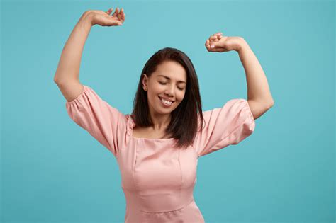 분홍색 드레스를 입은 어두운 머리를 가진 즐거운 행복한 미소 백인 여성은 손을 뻗고 아침 스트레칭을하고 사무실에서 졸린 느낌 푸른 배경에 고립 된 휴가를 꿈꾸고 있습니다