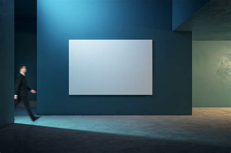 흐릿한 사업가가 콘크리트 바닥 모형이 있는 추상적인 홀 공간에서 어두운 벽 배경에 로고나 텍스트를 위한 장소가 있는 큰 흰색 빈 포스터를 걷고 있다 전시회에 대한 스톡 사진