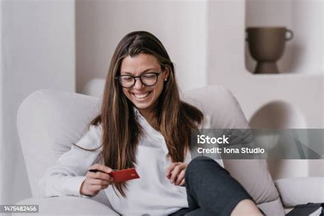 안경 흰색 셔츠 회색 바지를 입은 예쁜 갈색 머리 여자가 신용 카드를 들고 집에 앉아있는 아늑한 의자를 웃고 있습니다 사무실에서 매력적인 사업가는 큰 이익을 얻게되어
