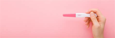젊은 성인 여성 손 밝은 분홍색 테이블 배경에 두 줄무늬와 임신 테스트를 들고 파스텔 색상 긍정적 인 결과 근접 촬영 관점 샷 와이드 배너 텍스트의 빈 장소입니다 맨 위