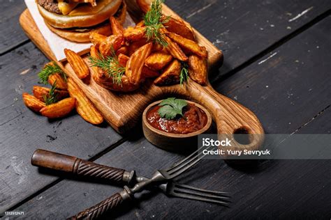 나무 도마에 튀긴 감자 웨지와 햄버거를 식욕을 돋우는 허브와 토마토 소스를 곁들인 구운 감자의 상당 부분을 차지합니다 패스트 푸드 포르 사진 0명에 대한 스톡 사진 및 기타