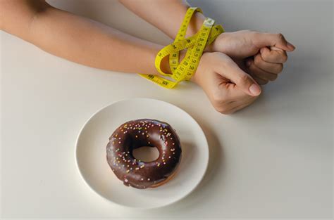 홈 오피스에서 부엌에서 책상에 노란색 측정 테이프와 맛있는 유리 초콜릿 도넛 접시로 묶여 여자 슬림 몸 손의 상단보기 정크 푸드 건강에 해로운 음식 다이어트 체중 감량 개념