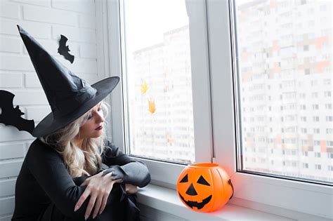 검은 마녀 의상을 입은 슬픈 금발과 얼굴 마스크가 창문 밖을 바라 봅니다 코비드로부터 할로윈 보호 준비 10월에 대한 스톡 사진