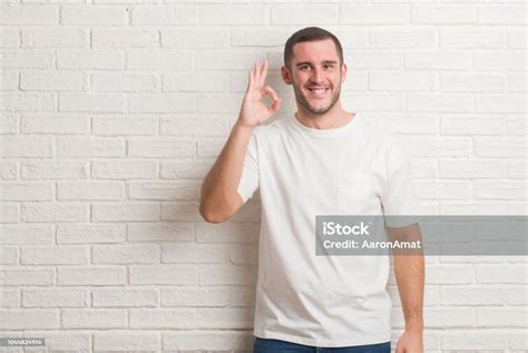 젊은 백인 남자 흰색 벽돌 벽 웃 고 긍정적인 손과 손가락으로 Ok 사인을 하 고 서 성공적인 식입니다 긍정적인 감정 표현에 대한 스톡 사진 및 기타 이미지 Istock