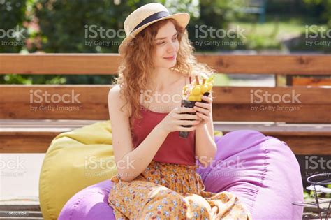 여우 곱슬 머리를 가진 젊은 여성의 야외 촬영 프레임없는 의자에 앉아 미소 옆으로 보고 그녀의 손에 큰 아이스크림을 들고 여름에