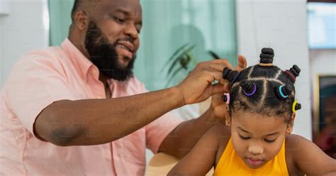 행복한 아프리카 가족 아버지는 거실에서 화려한 헤어 밴드로 어린 딸 머리를 묶고 있습니다 아빠와 아이 여자 아이는 집에서 뷰티