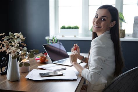 비즈니스 복장을 하고 웃는 듯한 여성이 조명이 환한 사무실의 책상에 앉아 노트북으로 작업하고 있습니다 그녀는 펜을 들고 사무용품 커피 한 잔 꽃이 든 꽃병에 둘러싸인 뒤를