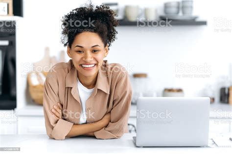 초상화 사랑스러운 긍정적 인 곱슬 머리 아프리카 계 미국인 젊은 여성 캐주얼 한 세련된 옷 여학생 또는 프리랜서 노트북 근처의 부엌에서 집에 서서 카메라를보고 행복하게 미소