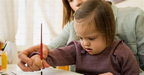 다운 증후군을 앓고있는 여자 아이가 직장의 테이블에 앉아있는 동안 종이에 페인트로 그림을 그리는 모습을 닫습니다 딸에게 그림을 그리는 것을 돕는 어머니 스톡 사진 사진