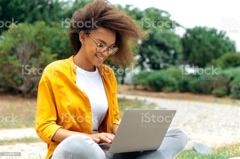 바쁜 성공적인 아프리카 계 미국인 곱슬 머리 젊은 여성 학생 소녀 프리랜서 세련된 캐주얼 한 옷을 입고 온라인 작업이나 공부를하는 동안 야외에서 노트북과 함께 앉아