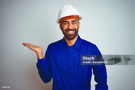 잘 생긴 인도 노동자 남자 유니폼과 헬멧 위에 고립 된 흰색 배경 미소 쾌활 한 제시 하 고 카메라를 보고 손바닥으로 가리키는