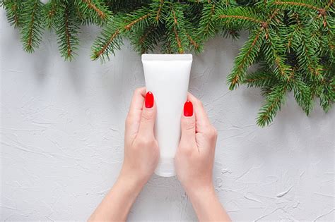 브랜드가 없는 흰색 스퀴즈 화장품 튜브 빨간 손톱과 회색 테이블에 크리스마스 전나무 나무의 가지와 여성의 손 천연 유기농 스파 화장품 컨셉 플랫레이 라이프 스타일 개념에