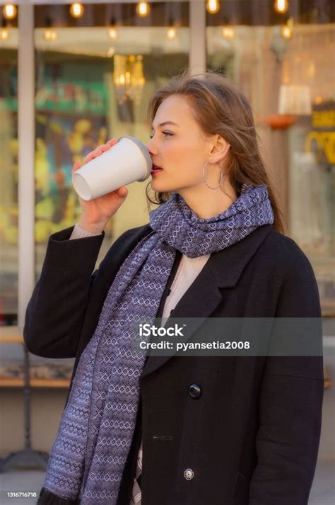 검은 코트와 스카프를 입은 젊은 미소 짓는 여성이 도시 쇼케이스 배경에서 생태재사용 가능한 컵과 커피를 들고 있습니다 친환경