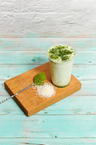 밝은 파란색 나무 테이블에 정물가에서 맛있는 차가운 여름 칵테일이 나무 스탠드에 말차와 코코넛 면도 숟가락과 함께 서 있습니다 코코넛 밀크에 말차와 밀크 쉐이크 0명에 대한