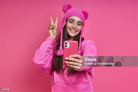 펑키 한 모자를 쓴 아름다운 젊은 여성이 셀카를 만들고 분홍색 배경에 몸짓을합니다 즐거움에 대한 스톡 사진 및 기타 이미지