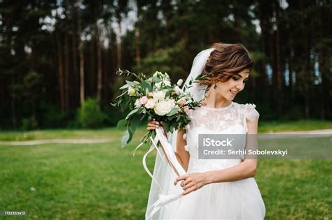 우아한 드레스에 젊은 아름 다운 신부는 자연에서 리본으로 분홍색 꽃과 녹색의 꽃다발을 들고 숲 근처 필드에 서 있다 야외 결혼식 후 개념에 대한 스톡 사진 및 기타 이미지