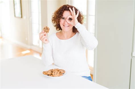 집에서 초콜릿 칩 쿠키를 먹는 시니어 여성 행복한 얼굴로 손가락을 통해 보고 있는 눈으로 확인 사인을 하고 미소 짓고 있습니다 가정 생활에 대한 스톡 사진 및 기타 이미지