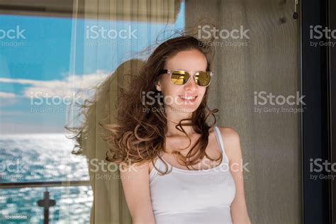 선글라스를 착용한 아름다운 젊은 로맨틱 한 긴 머리 의 여성은 발코니의 파노라마 창문 옆에 서서 미소를 지으며 바다 풍경을 보고 있습니다 갈색 머리에 대한 스톡 사진 및