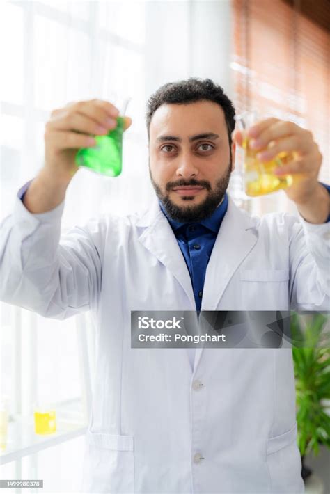 플라스크를 들고 있는 실험복을 입은 백인 과학자 남자는 실험실에서 녹색과 노란색 용액을 들고 있다 실험을 하기 위하여 학생을 위한 해결책 그리고 몇몇 시약 준비 Stem