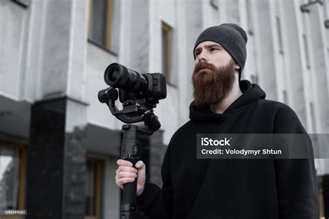 3축 짐벌 안정기에 전문 카메라를 들고 있는 검은 후드를 입은 수염 전문 비디오 그래퍼 전문 영화 카메라로 훌륭한 비디오를 만드는 영화 제작자 촬영 베트남에 대한 스톡 사진