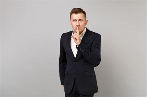엄격한 젊은 사업가 자장을 말하는 고전적인 검은 양복 셔츠에 회색 배경에 고립 입술 쉿 제스처에 손가락으로 조용할 성과 경력 부 비즈니스 개념입니다 복사 공간을 비웃는 다