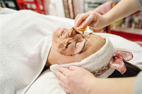 스파 살롱 미용실에서 커피 얼굴 트리트먼트를 가진 젊은 여성은 침대 머리에 누워 있는 미용실 미용 치료에서 소녀의 얼굴에 벗겨진 스폰지 마스크를 제거합니다 건강관리와 의술에