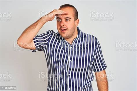 고립 된 흰색 배경 위에 서있는 파란색 줄무늬 셔츠를 입은 청년은 매우 행복하고 머리 위로 손으로 멀리 보고 미소 개념 검색