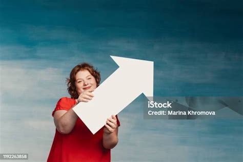 빨간 티셔츠와 청바지에 예쁜 빨간 머리 통통 한 여자는 파란색 배경에 측면에 흰색 위로 화살표를 보유하고 있습니다 T 셔츠에 대한 스톡 사진 및 기타 이미지 Istock