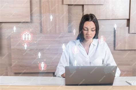 사무실 배경 위에 Hr 소셜 미디어 홀로그램 아이콘을 컨설팅하는 국제 비즈니스 컨설팅을 위해 새 직원을 고용하기 위해 노트북으로 작업하는 직장에서 흰 셔츠를 입은 매력적인