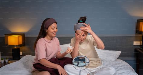 웃는 아시아 여자 친구들은 침대에 앉아 집에서 함께 얼굴에 스킨 케어 페이셜 마스크를 바르고 있다 여성 게이 커플은 긴장을 풀고 행복과 함께 뷰티 페이셜 트리트먼트를