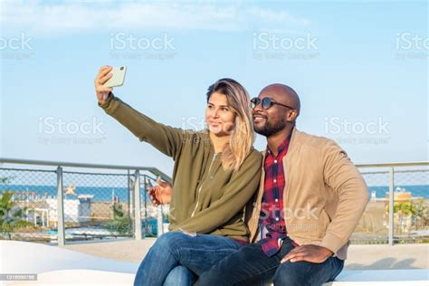 봄 방학 동안 공원에서 벤치에 앉아 셀카를 찍는 다민족 볼트 남자와 백인 여성의 행복한 그룹미소 는 소년과 소녀의 인종 간 커플야외에서 자화상 사진을 찍고햇빛이 그들을